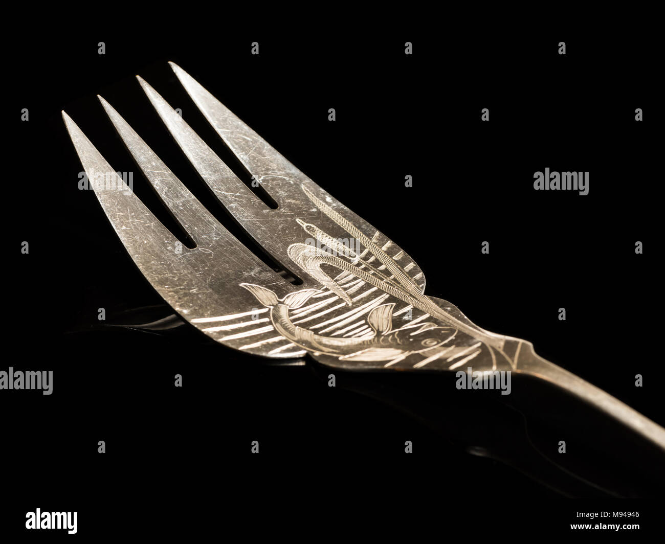 Fish fork made of silver with engravings on a black reflective surface ...