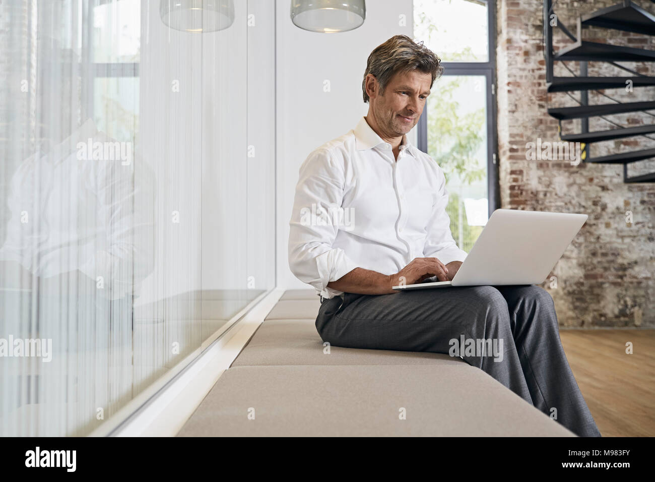 Businessman sitting on bench in modern office using laptop Stock Photo ...