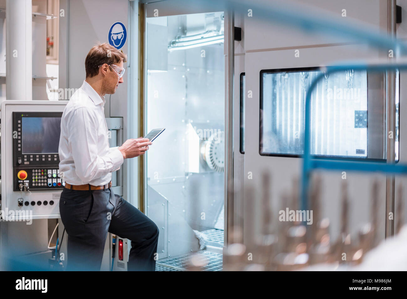 Businessman at machine in factory wearing safety goggles holding laptop ...