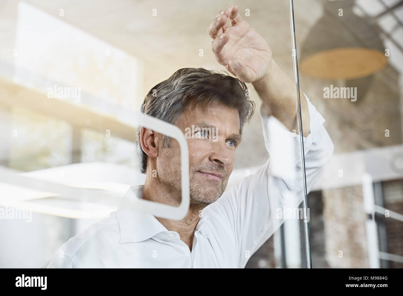 Portrait of mature businessman leaning against glass wall in office ...