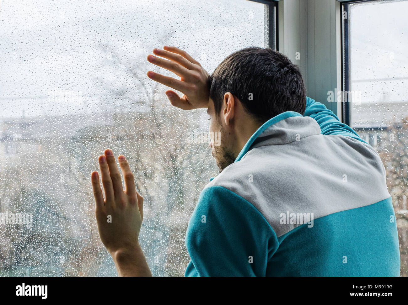 Depressed young man upset with bad news looking through rainy window ...