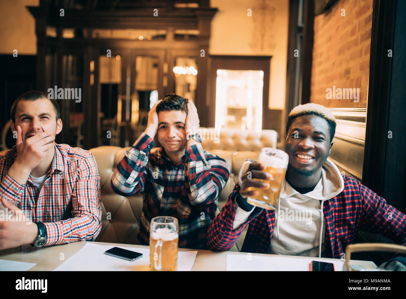 Watching TV in bar. Two happy young men drinking beer and gesturing ...