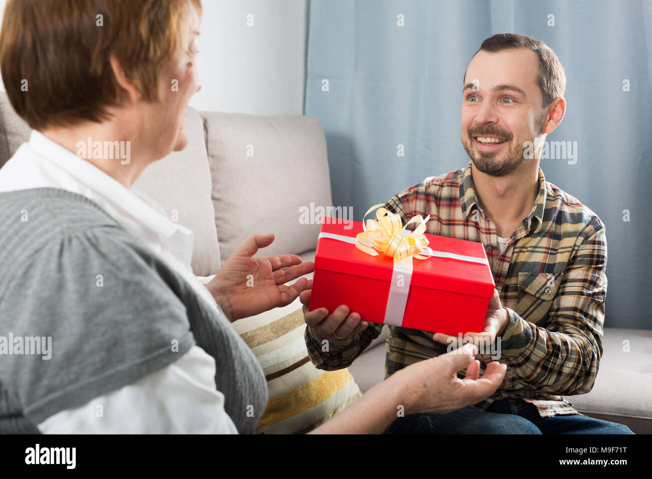 Adult son giving present to aged mother during evening together at home ...