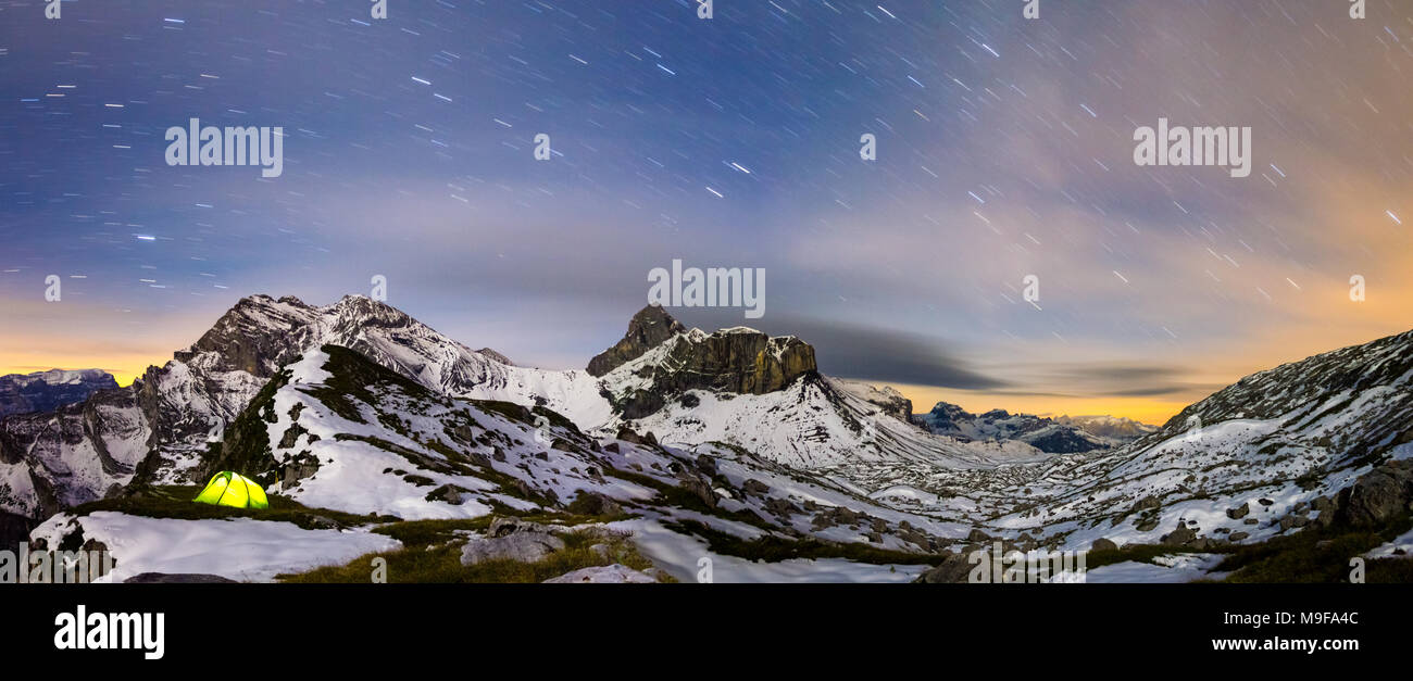 Panorama of illuminated tent under starry night sky in snowy alpine mountains. Alps, Switzerland ...