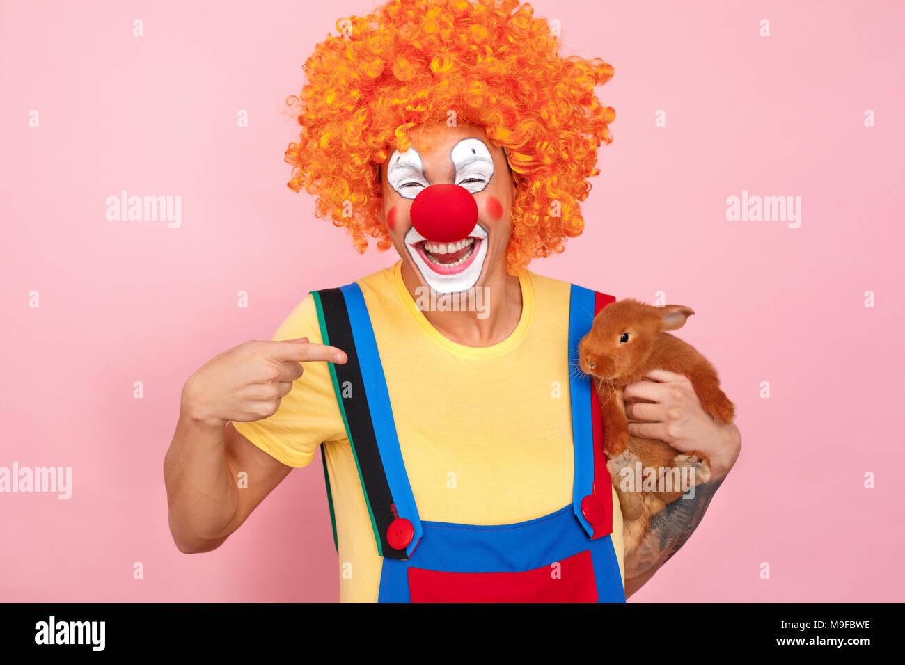 Man in a red wig, overalls and makeup holding small rabbit and pointing ...