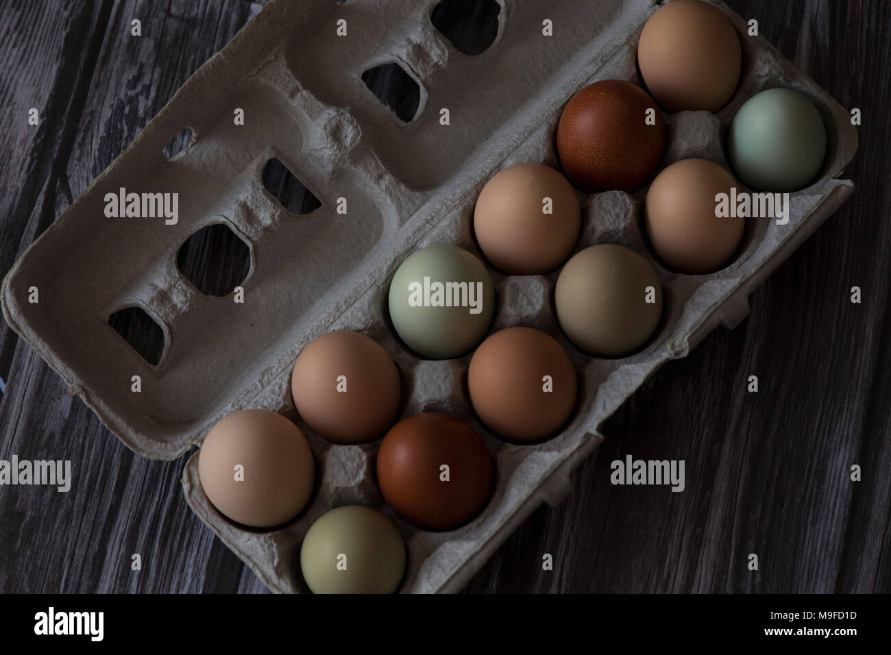 A carton of a dozen farm fresh eggs in multiple colors, on a wooden ...