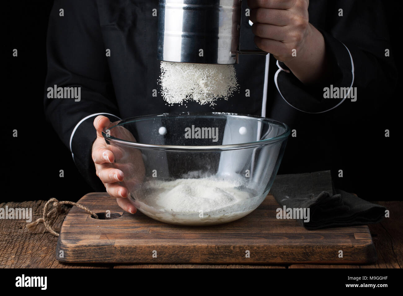 Chef hands pouring flour powder on raw dough using sieve on a black ...