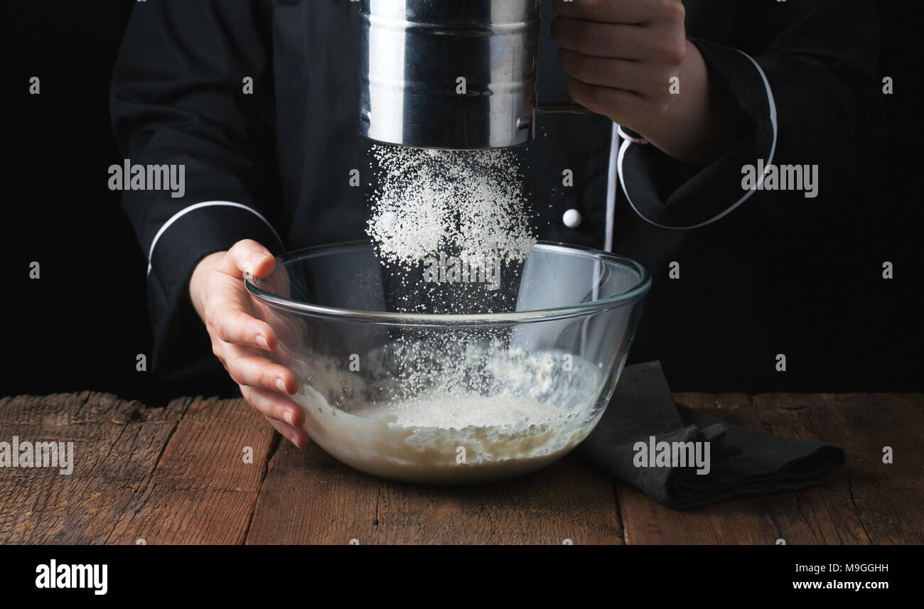 Chef hands pouring flour powder on raw dough using sieve on a black ...
