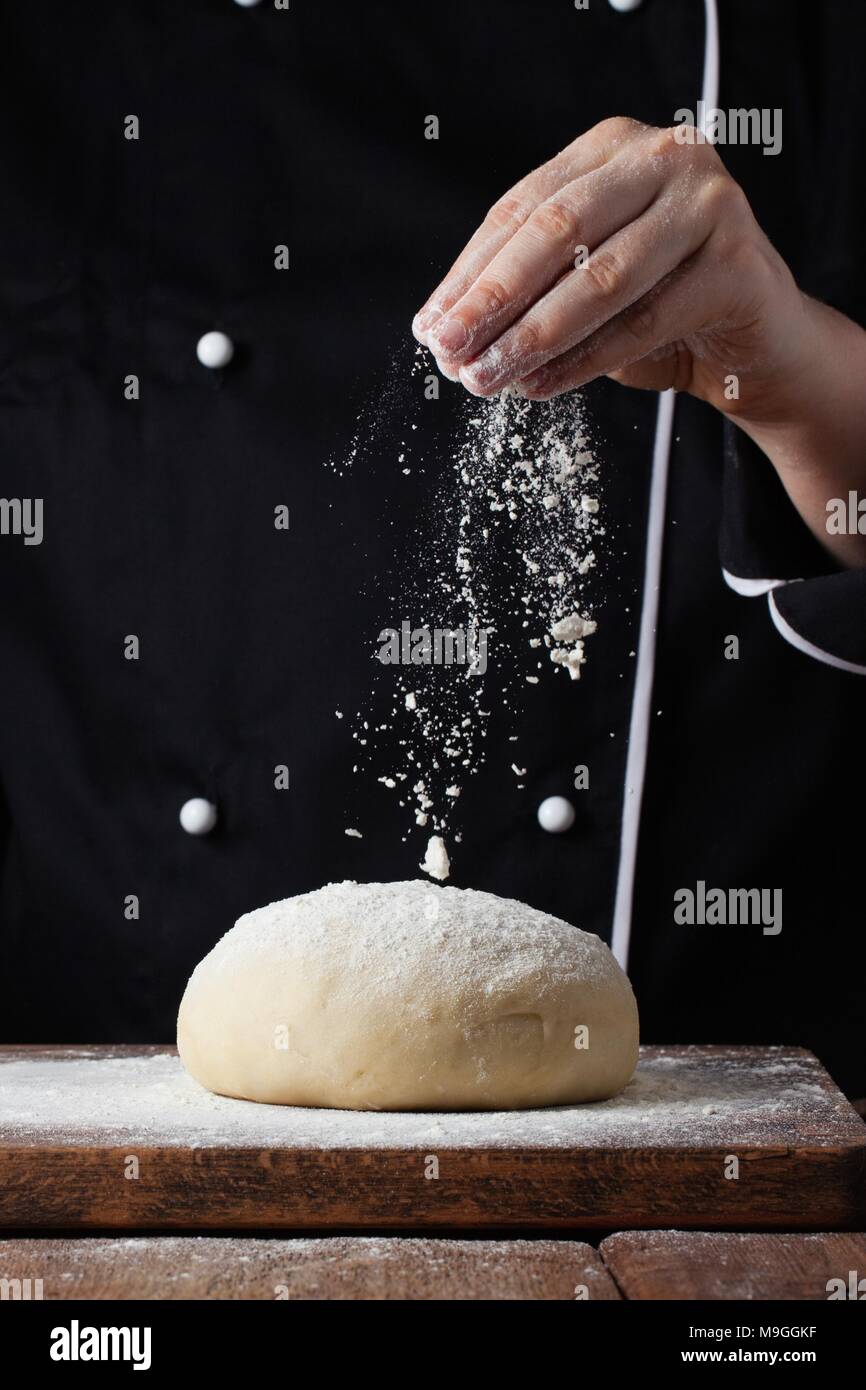 Chef hands pouring flour powder on raw dough using sieve on a black ...