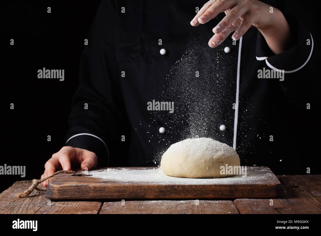 Chef hands pouring flour powder on raw dough using sieve on a black ...