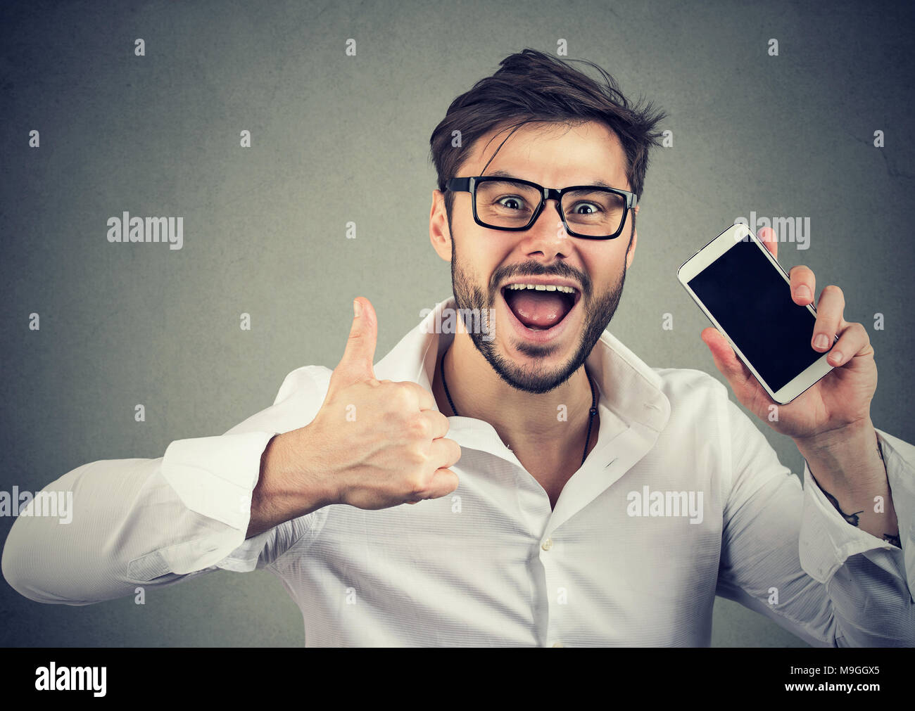 Super excited young man in eyeglasses showing smartphone and thumb up ...