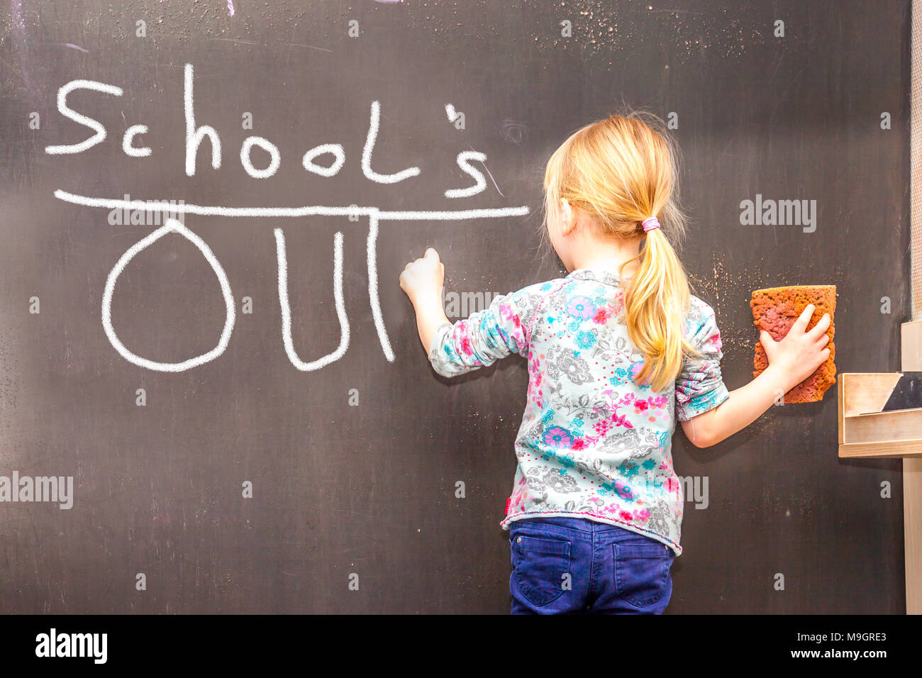 Cute little girl writing Schools's out on chalkboard in a classroom ...