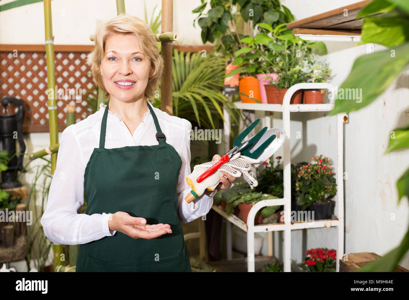 Happy mature woman florist in apron holding trowel and small rake for ...