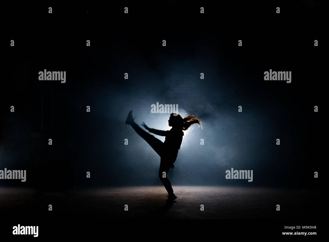 young dancer with long hair raising up her leg on dark stage. flexible ...