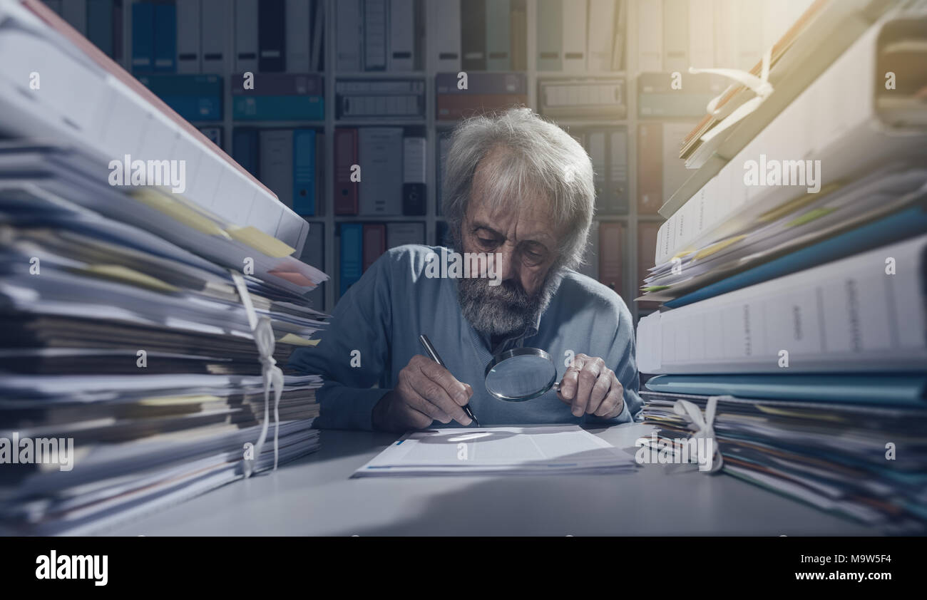 Senior researcher using a magnifying glass and writing, his desk is ...