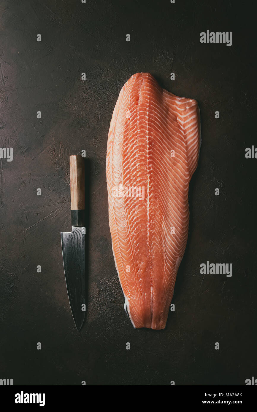 Whole raw uncooked salmon fillet with chef's knife over dark brown ...