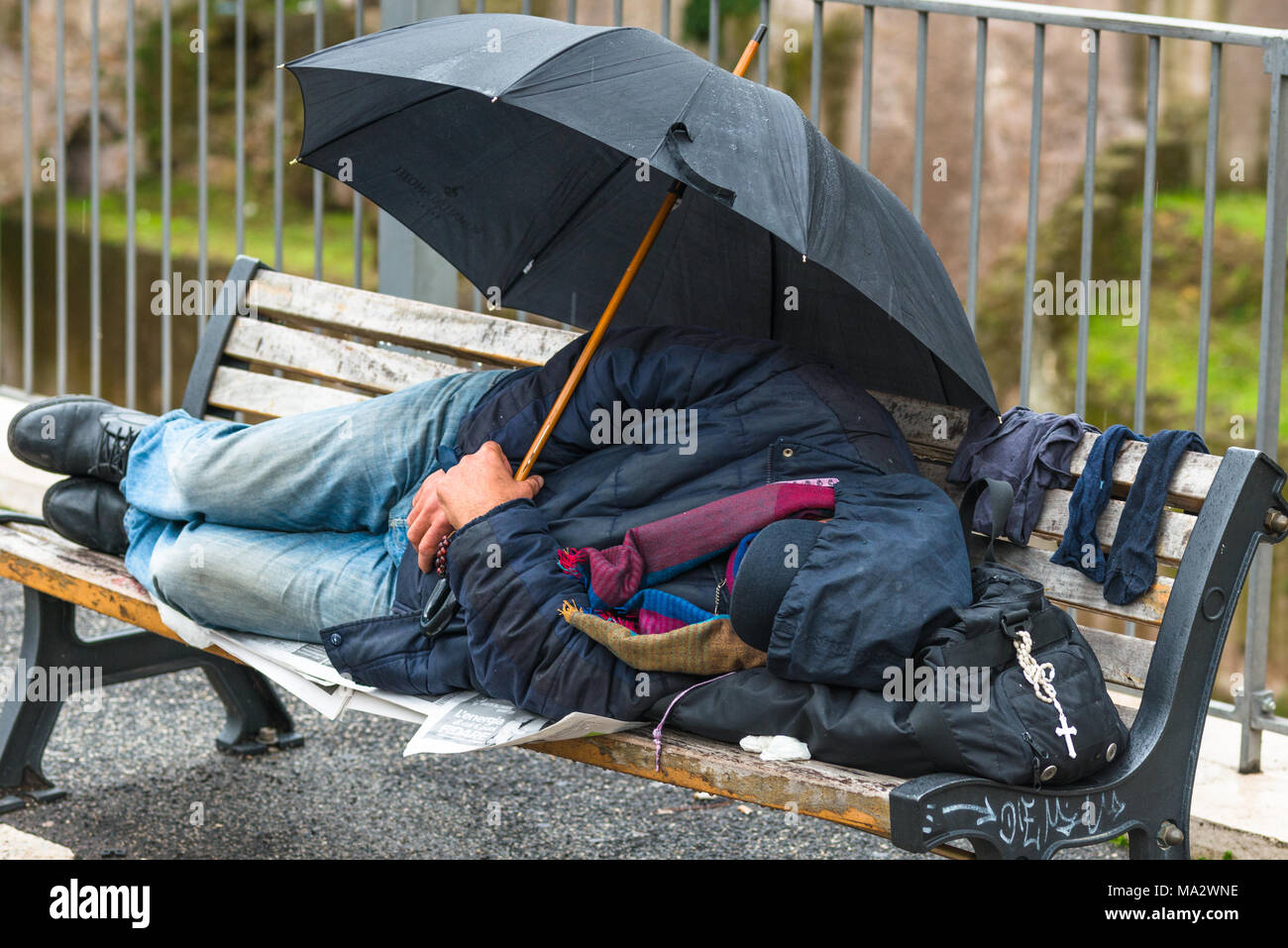 A homeless man in the rain on a bench with an umbrella on Via dei Fori ...