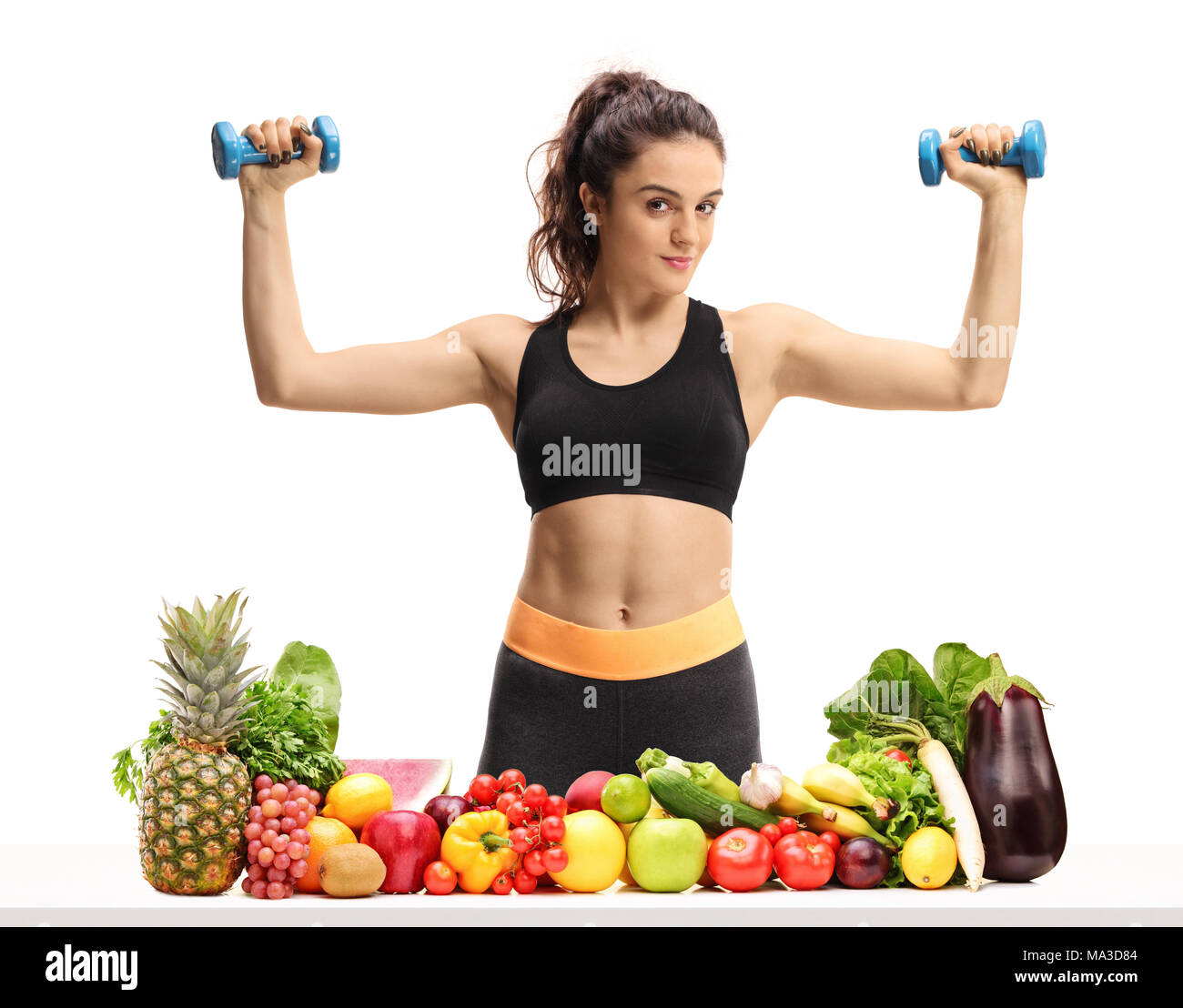 Fitness woman exercising with dumbbells behind a table with vegetables ...