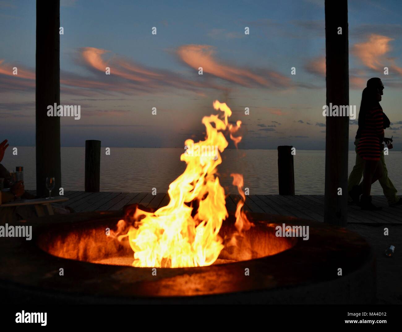 Flickering flames in outdoor fire pit along pier with couple walking by ...