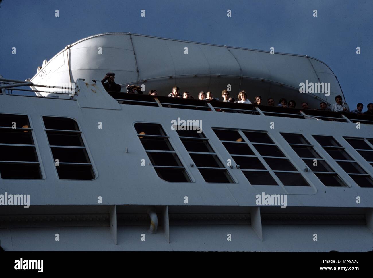 Passengers look over the rail of a cruise ship, with a large tender or ...