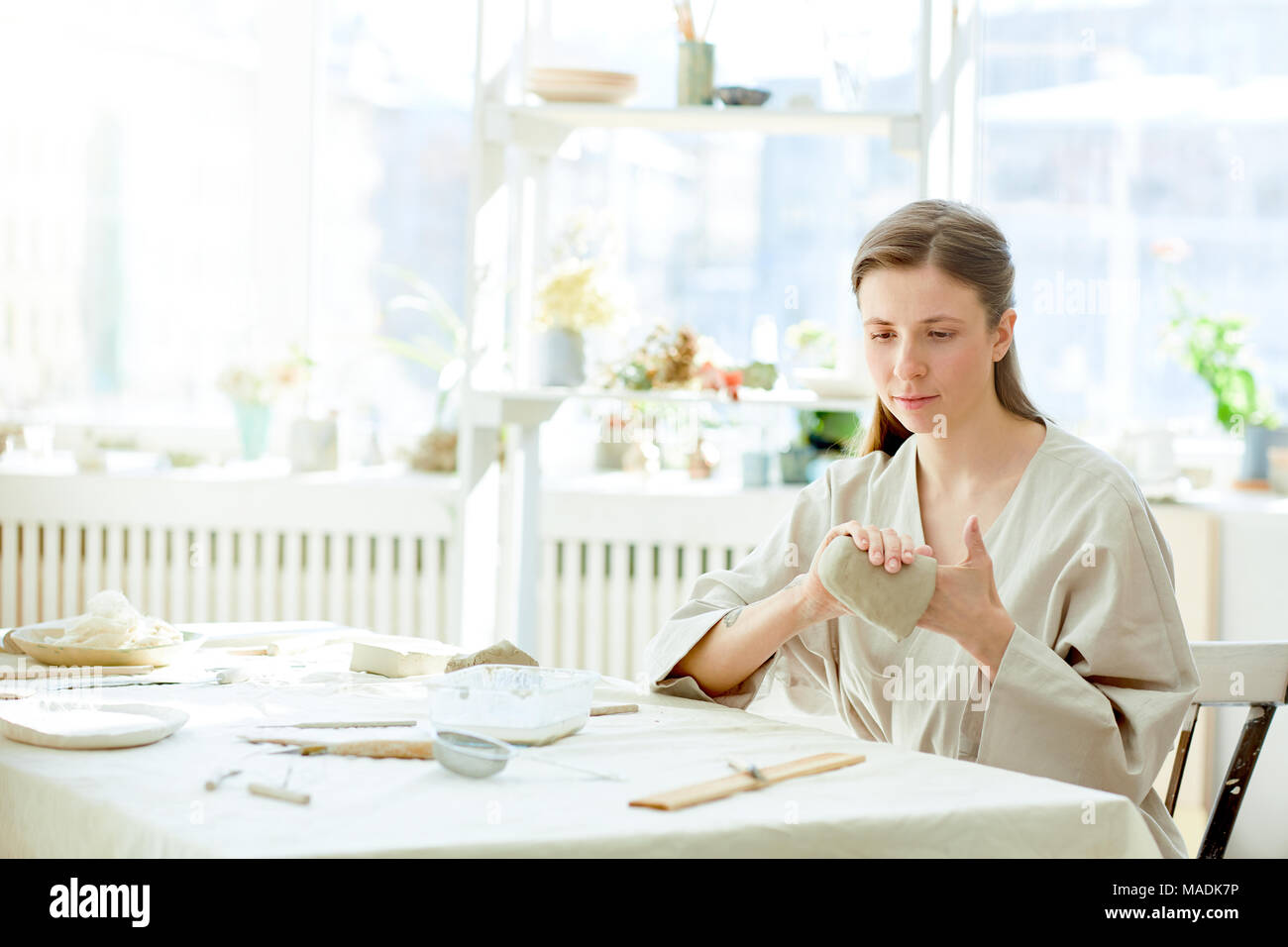 Young creative woman in grey robe sitting by table and making mug from ...