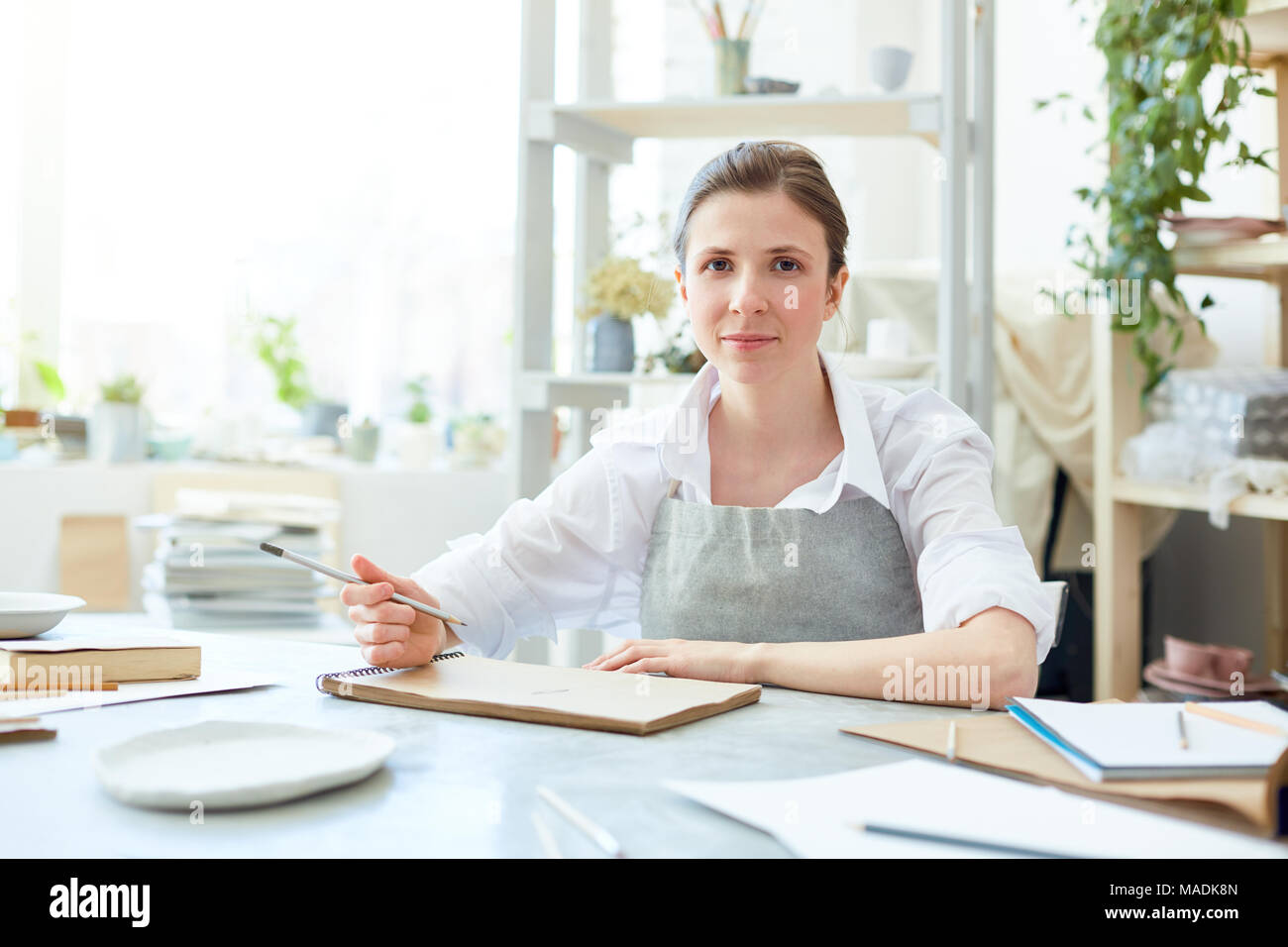 Young creative woman in white shirt and grey apron sitting by table and ...