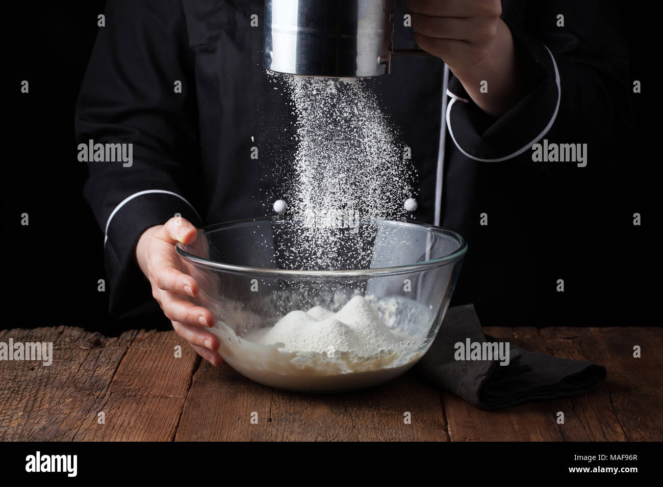 Chef hands pouring flour powder on raw dough using sieve on a black ...