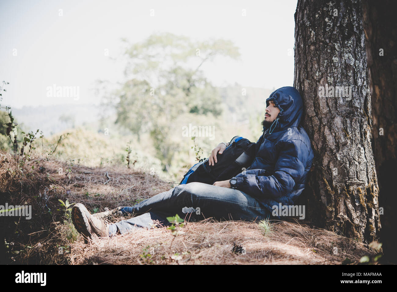 Portrait of young beard man sitting alone by a tree with backpack ...