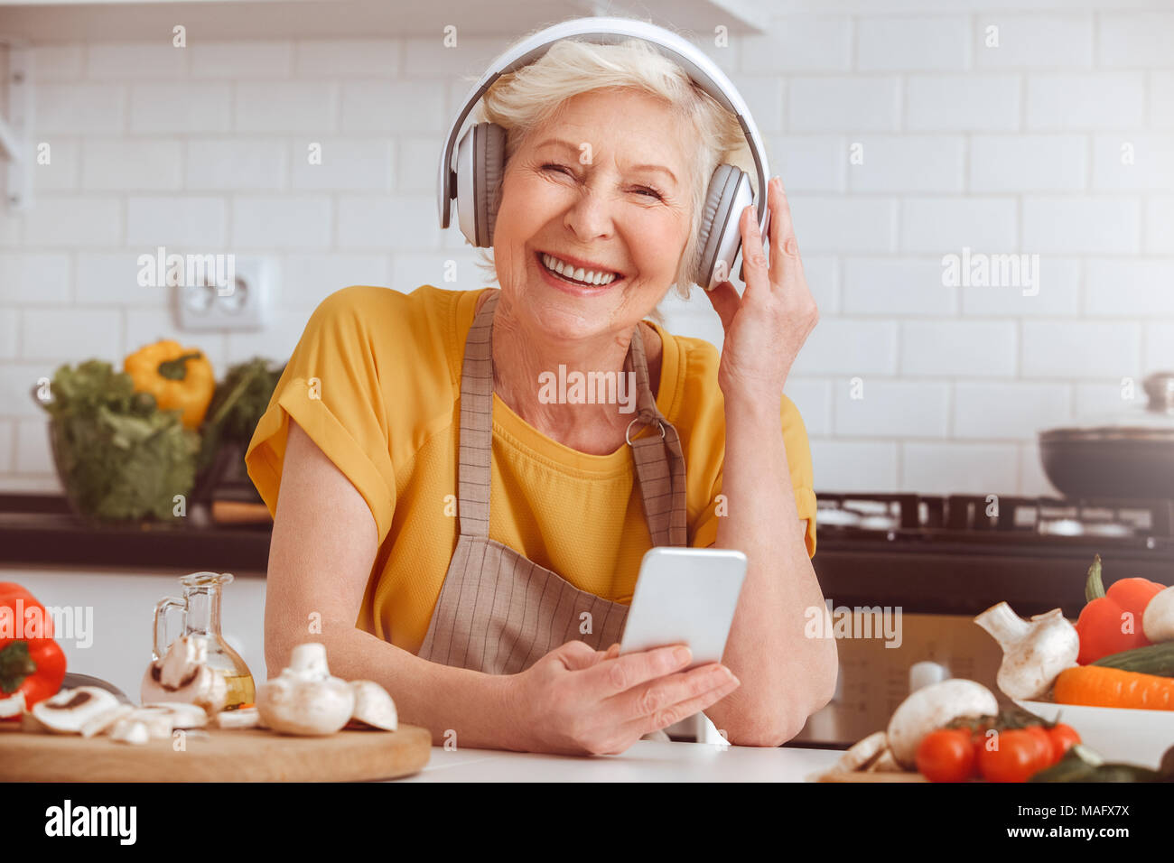 Older woman cooking, listen to music in mobile, toothy smiling. Indoor ...