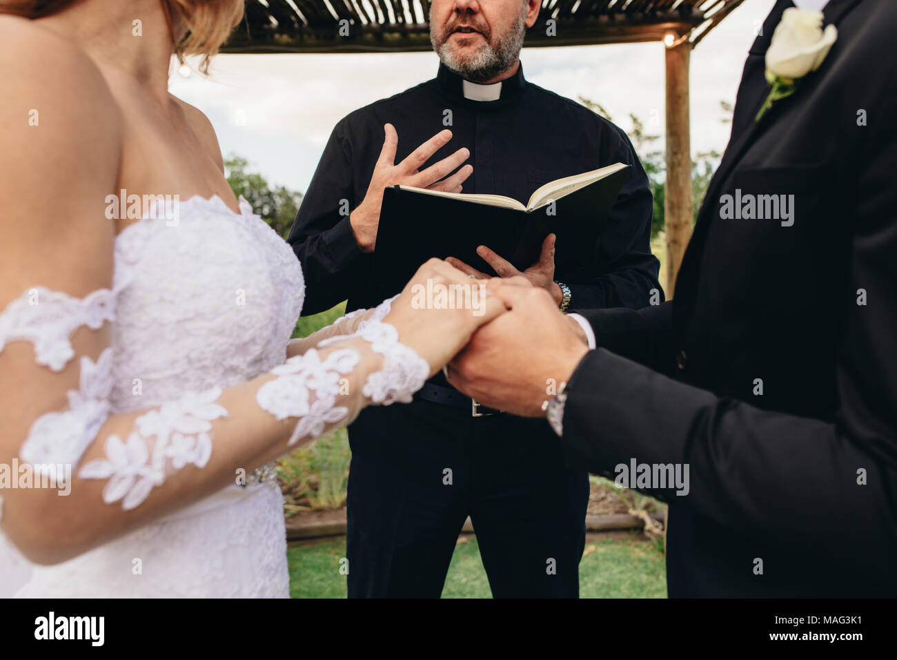 Bride and groom holding hands in presence of priest reading prayers ...