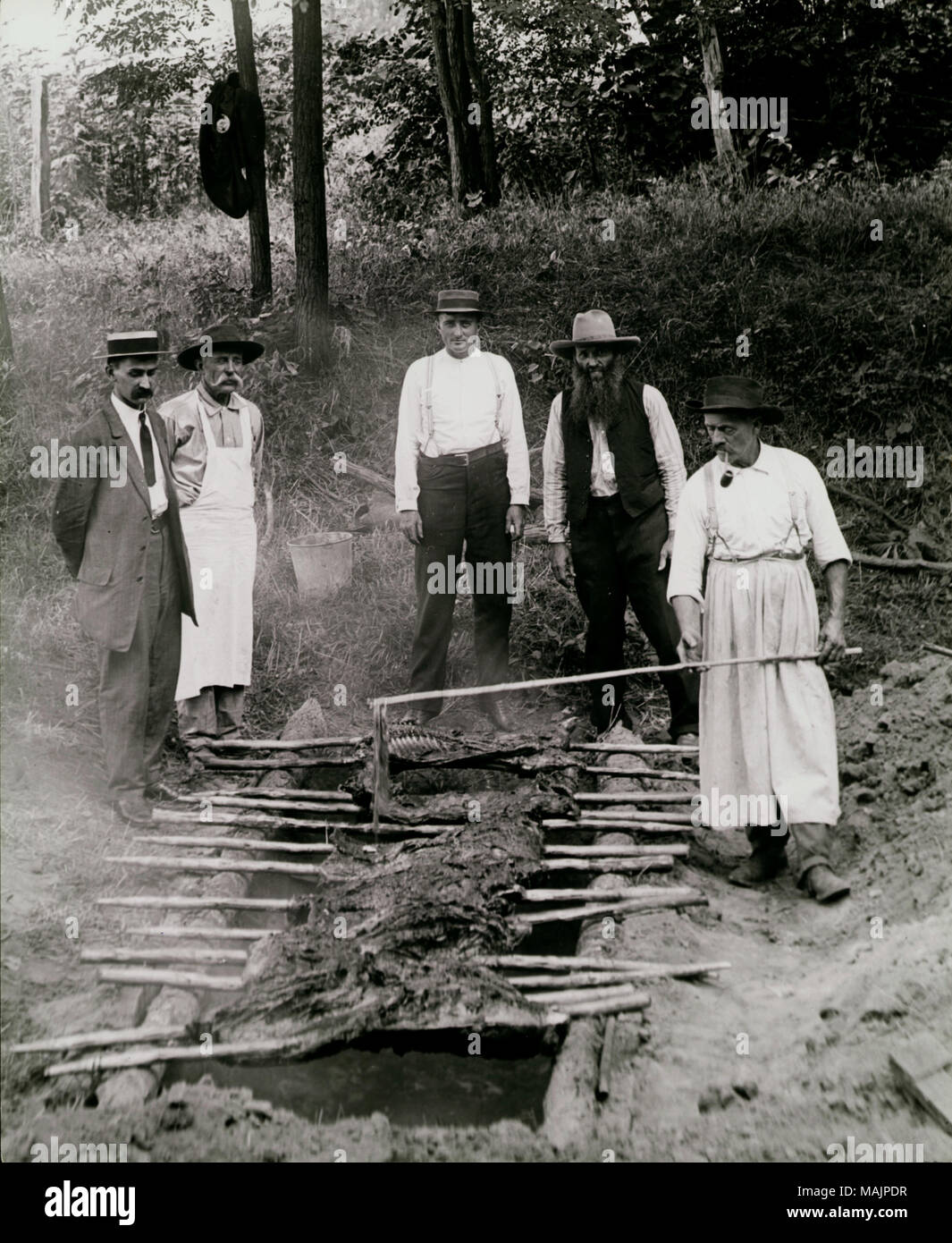 Title: Five men cooking spitted meat on pit barbeque in rural setting ...