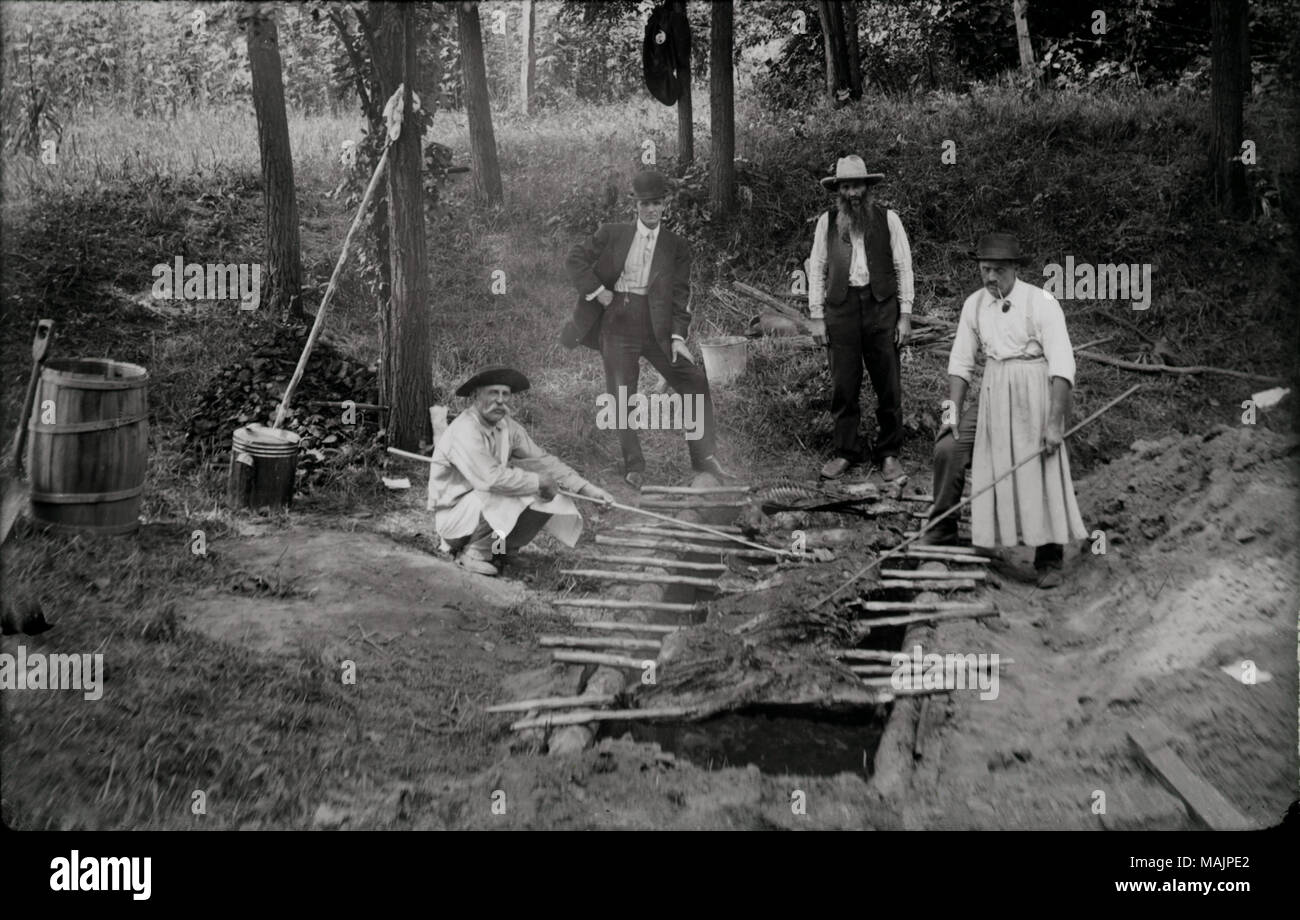Title: Four men cooking spitted meat on pit barbeque in rural setting ...