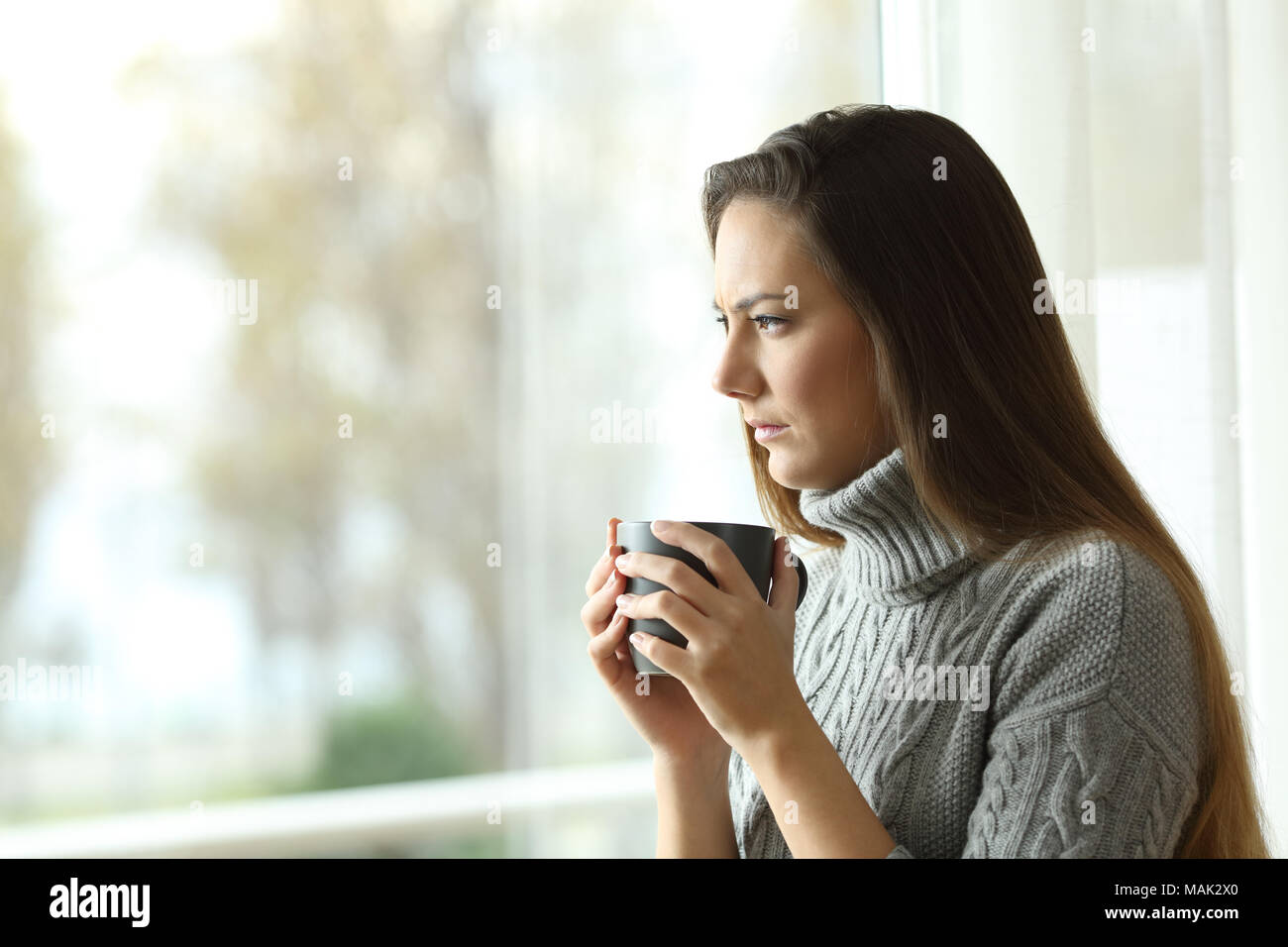 Angry woman looking through a window and holding a mug at home in ...
