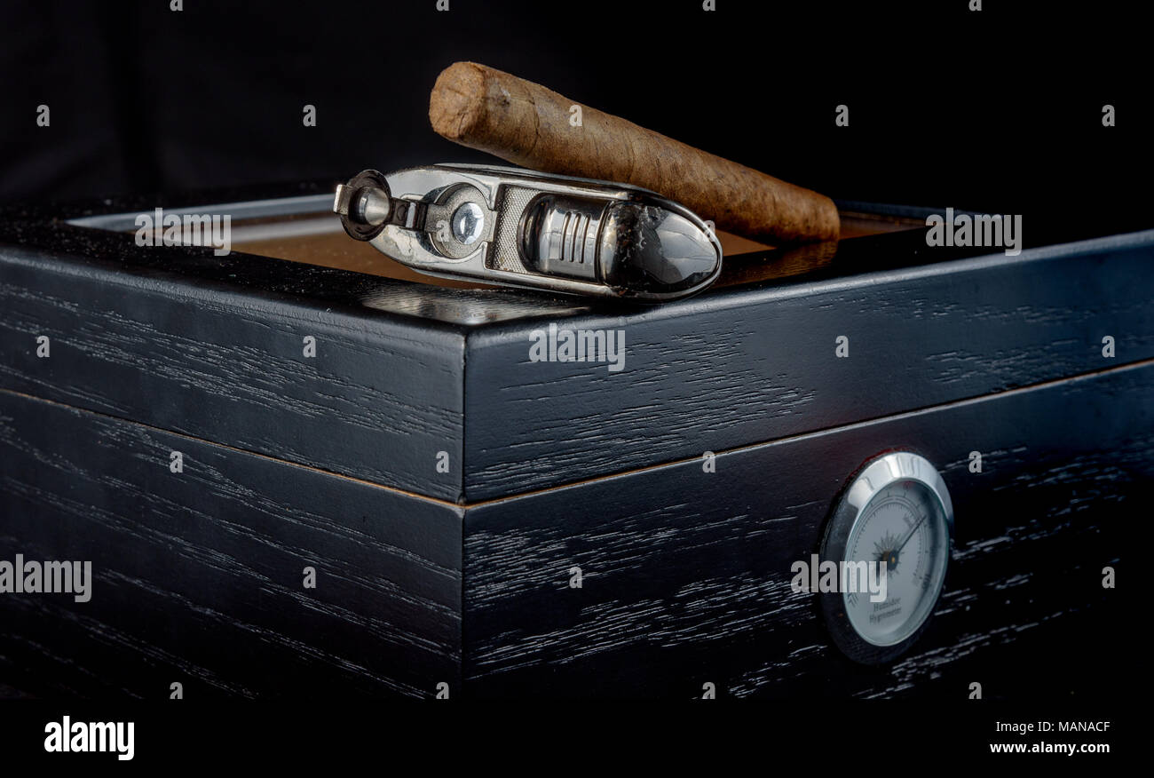 A rich smelling cigar on a black humidor next to a cigar punch torch ...