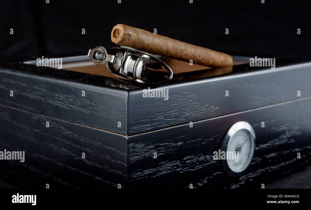 A rich smelling cigar on a black humidor next to a cigar punch torch ...