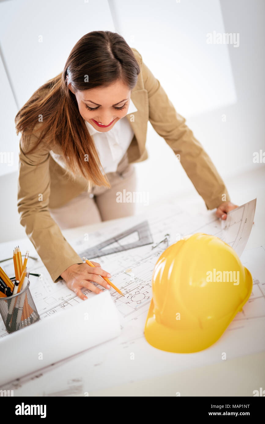 Smiling young woman architect constructor analyzing blueprint at desk ...