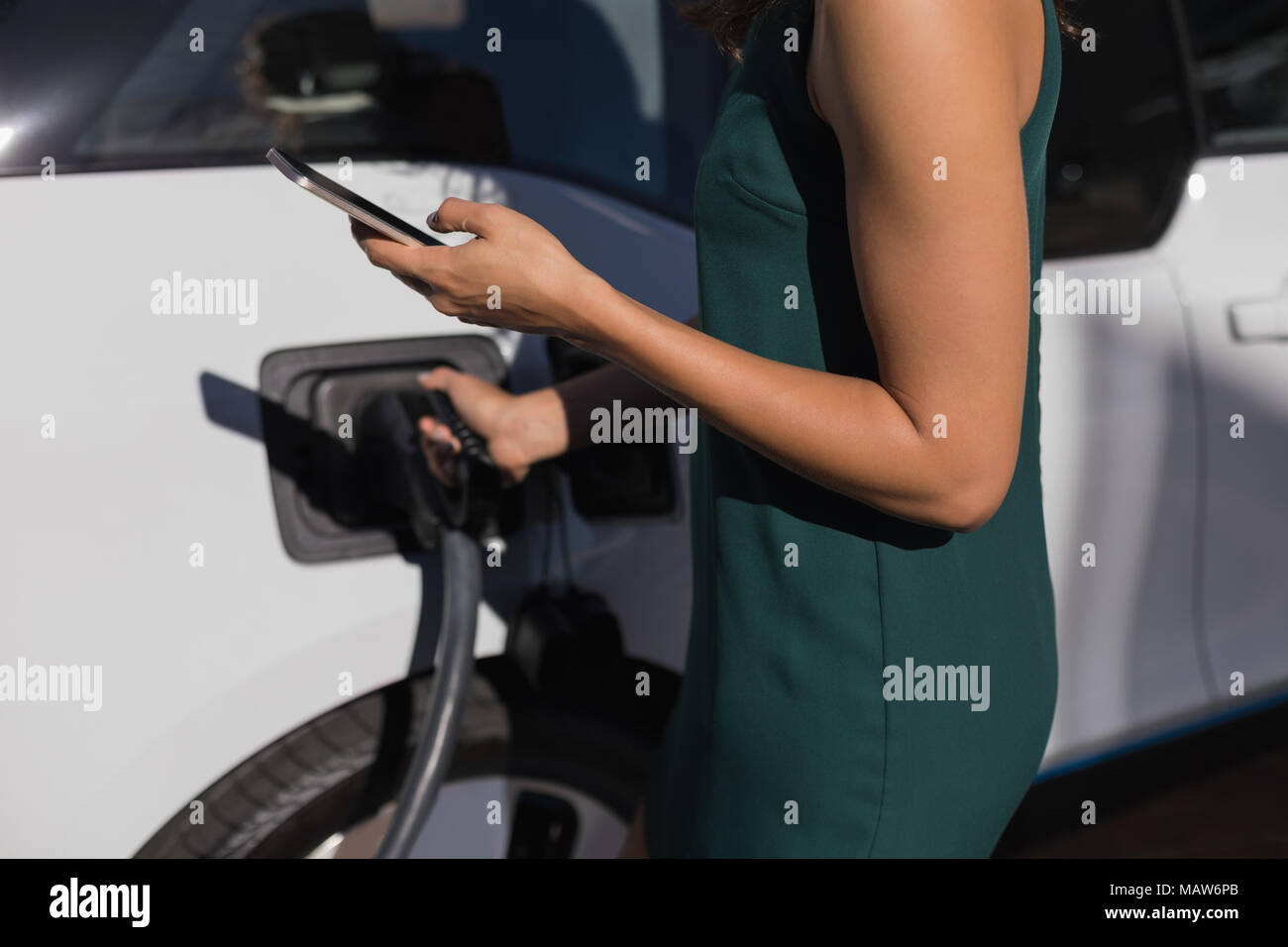 Woman using mobile phone while charging electric car at charging ...