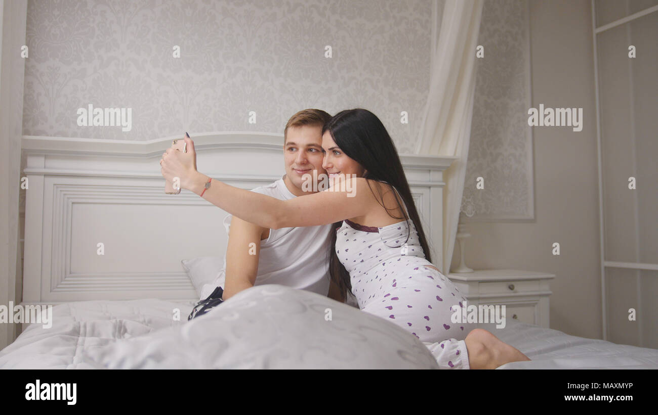 Happy cheerful loving couple making selfie in bed, young attractive guy ...