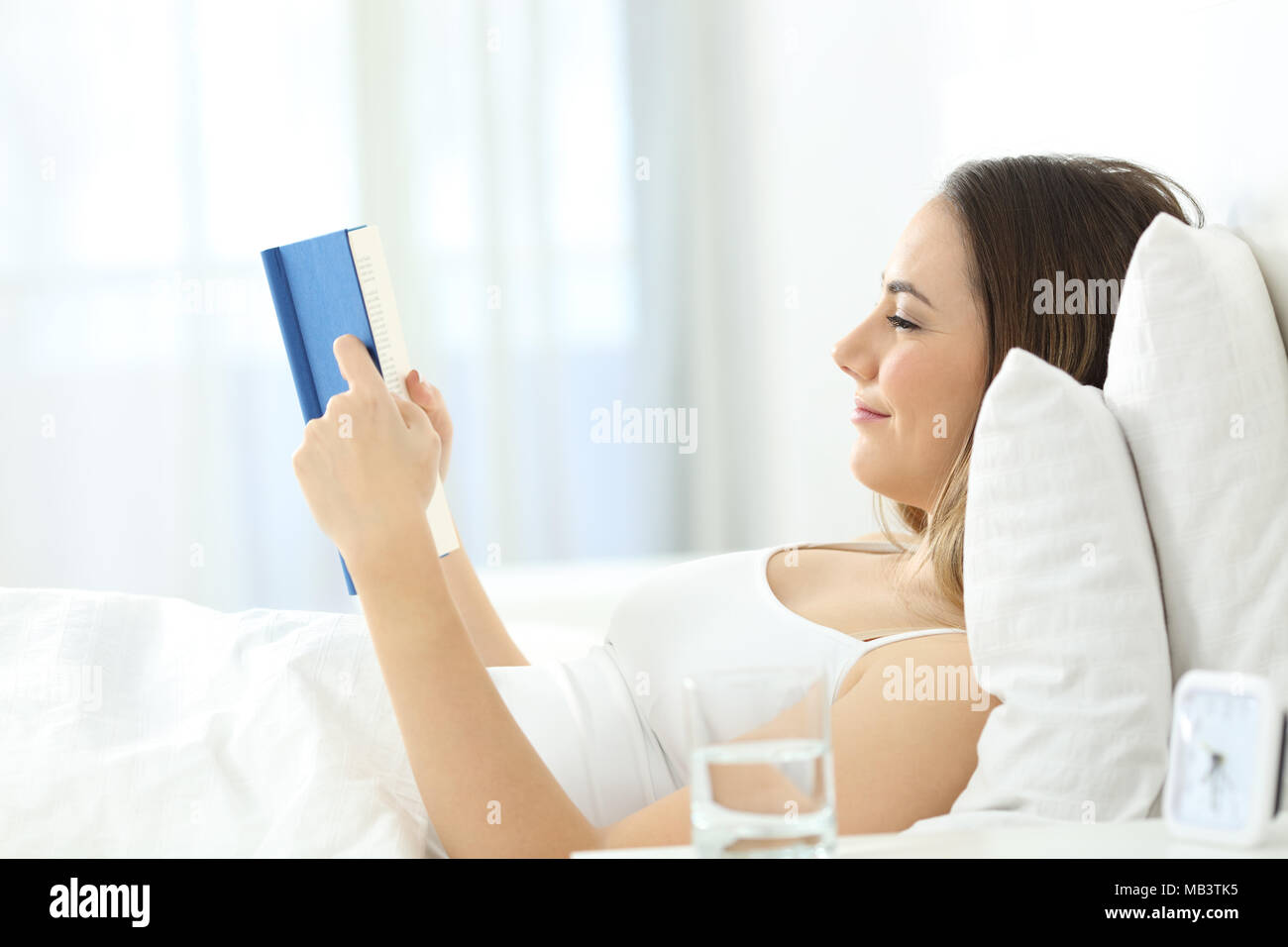 Profile of a satisfied woman reading a paper book on the bed at home ...