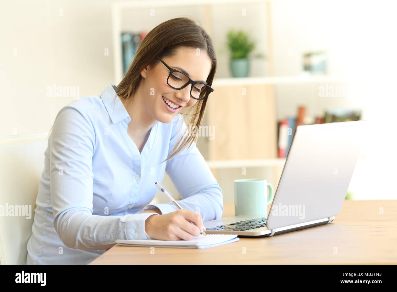 Happy woman wearing eyeglasses writing in a notebook on a table at home ...