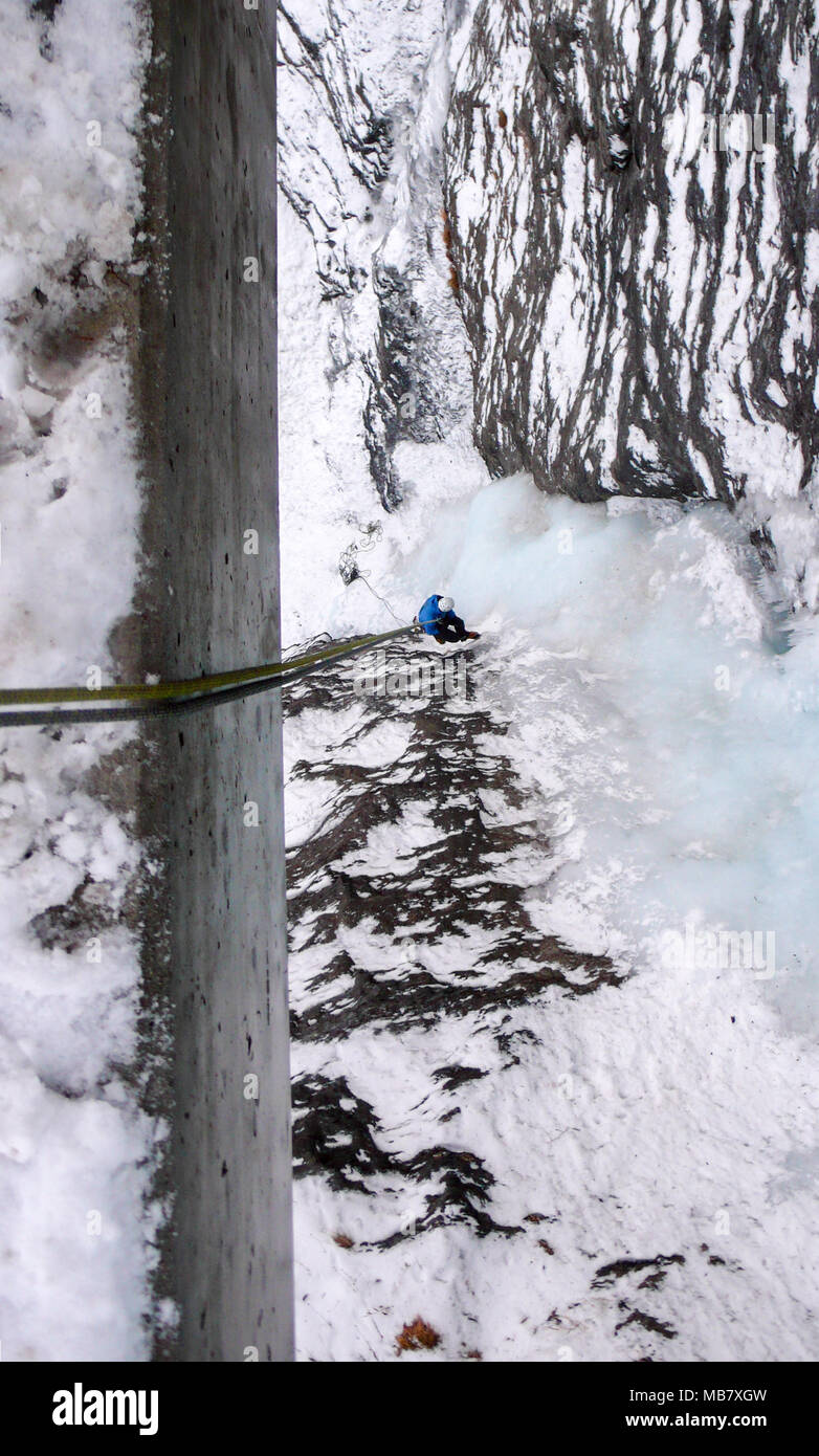 male ice climber in a blue jacket rappelling a from a bridge railing ...