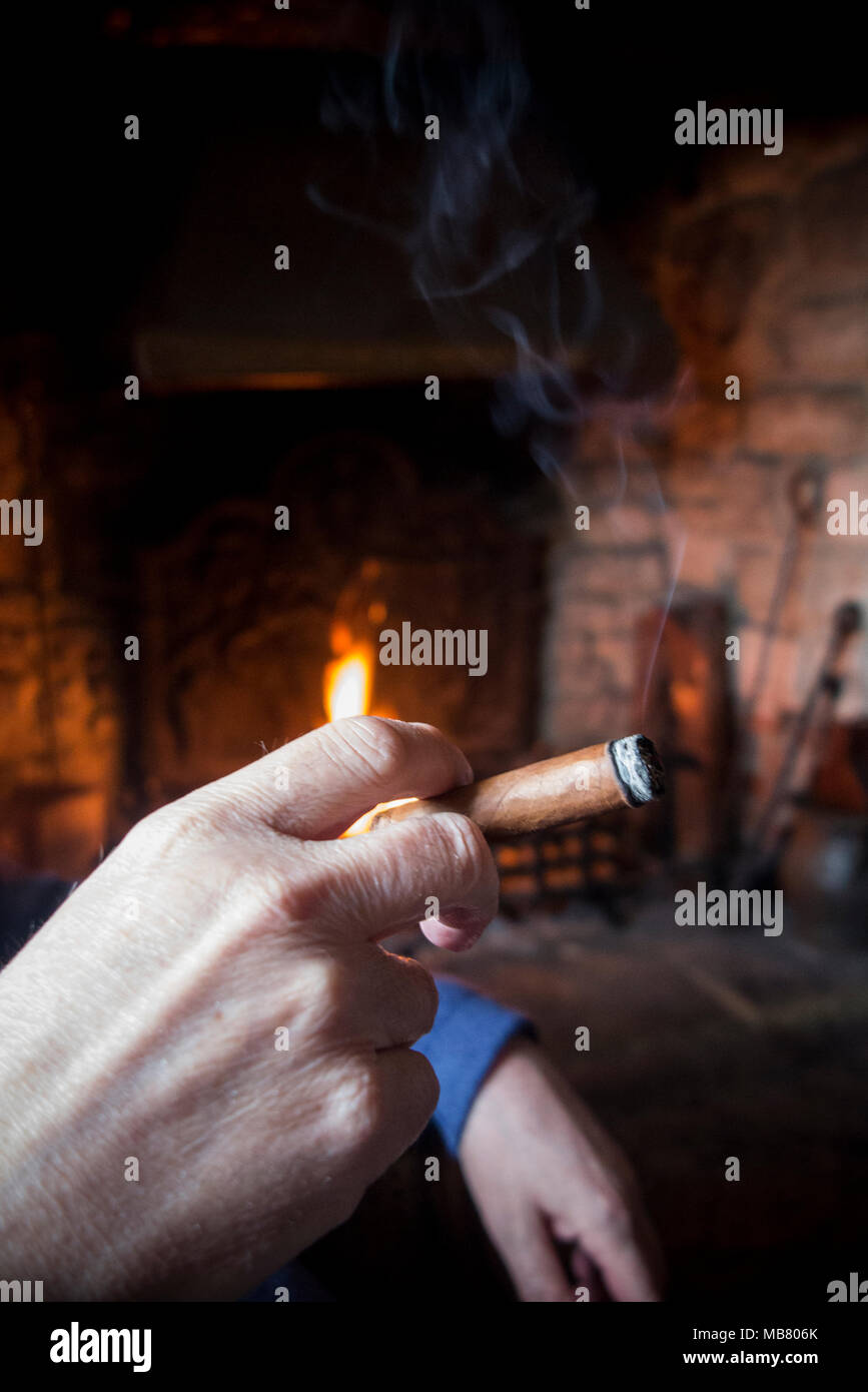 A man smokes a cigar in front of a roaring fire in a country house ...