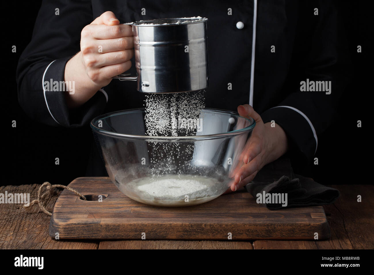 Chef hands pouring flour powder on raw dough using sieve on a black ...