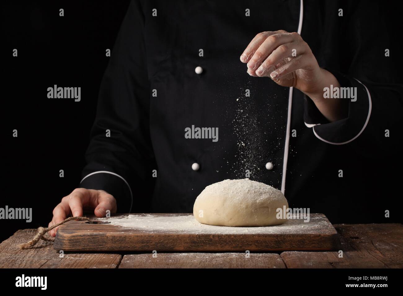Chef hands pouring flour powder on raw dough using sieve on a black ...