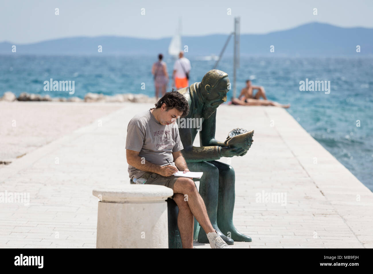 Man writing in a notebook sitting besides the statue of a scientist ...
