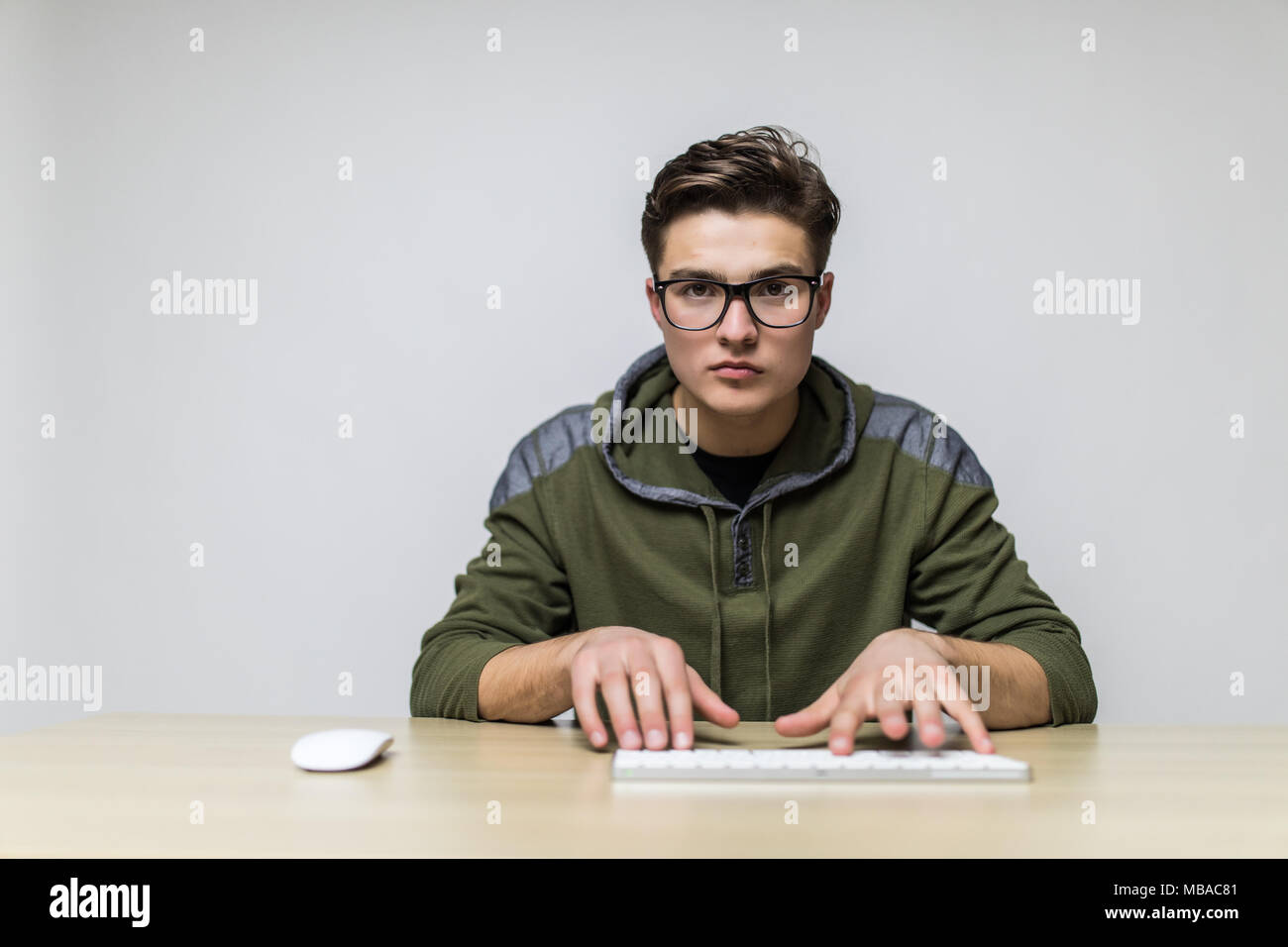 A young hacker with glasses dressed in casual clothes sitting at a desk ...