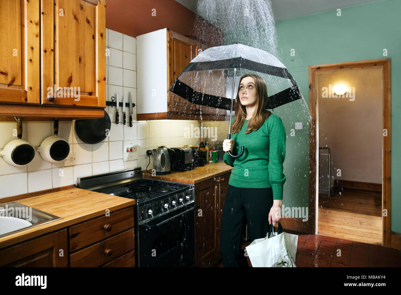 Woman standing in kitchen with umbrella under shower of rain, indoors ...