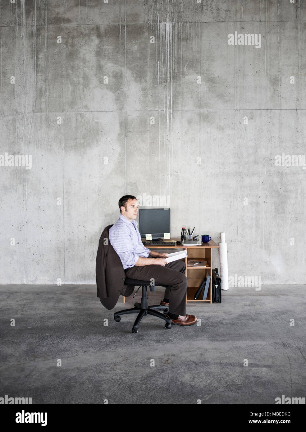 Business man sitting at a small desk in a large empty raw office space ...
