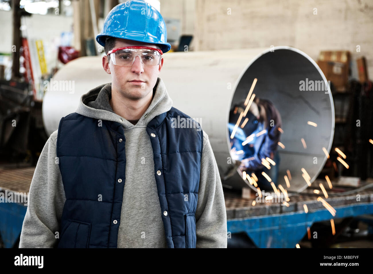 A young caucasian factory worker wearing a hard hat in a sheet metal ...