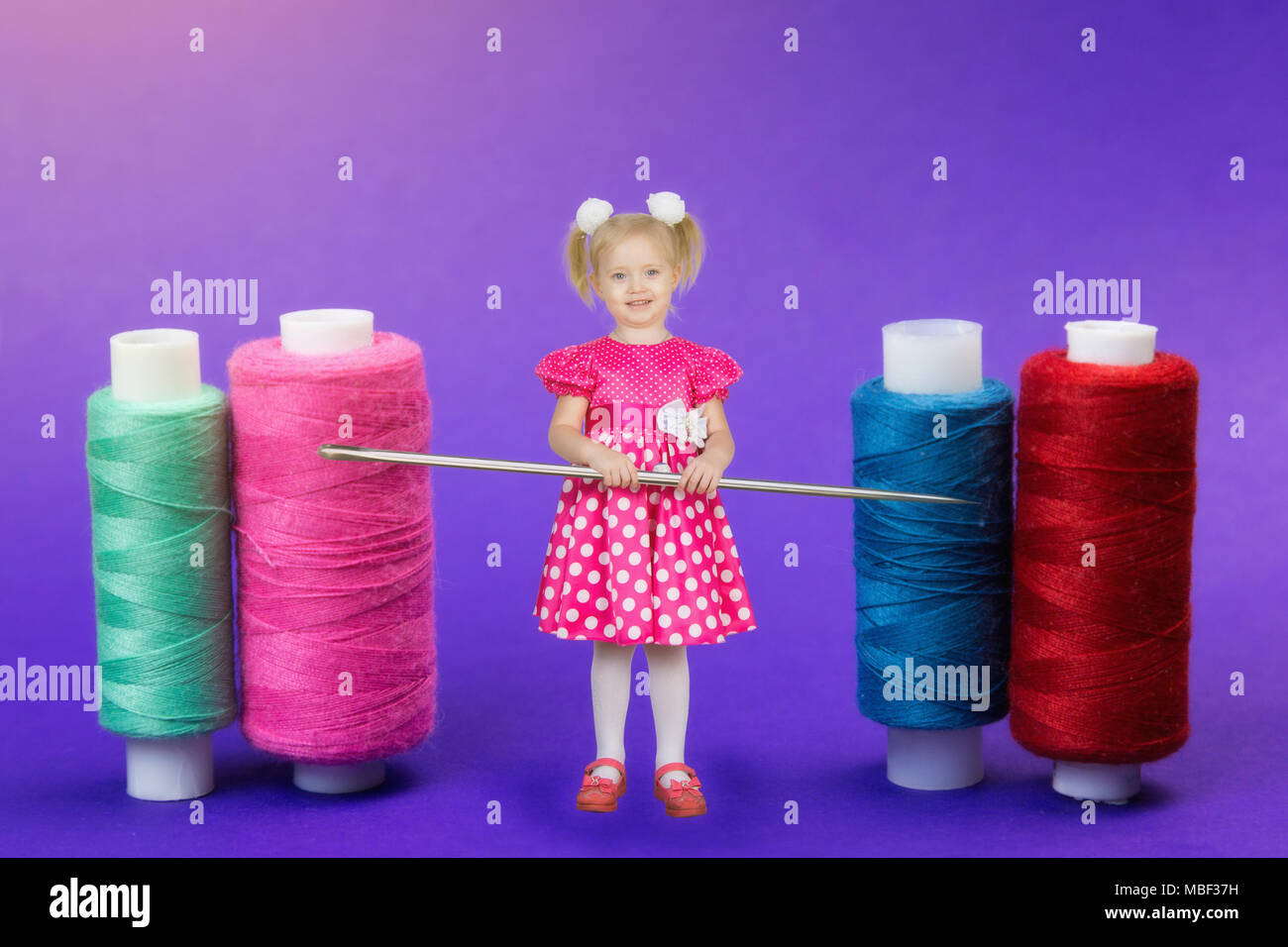 Blonde girl 3 years old in a red dress is holding a large needle for ...
